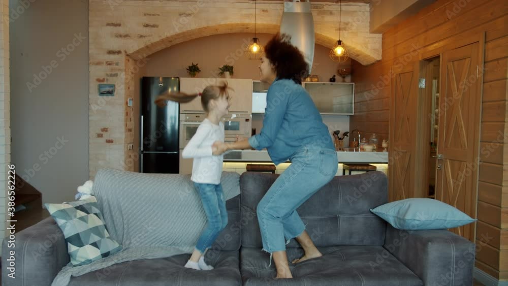 Playful girl and Afro-American woman babysitter are jumping on sofa in ...