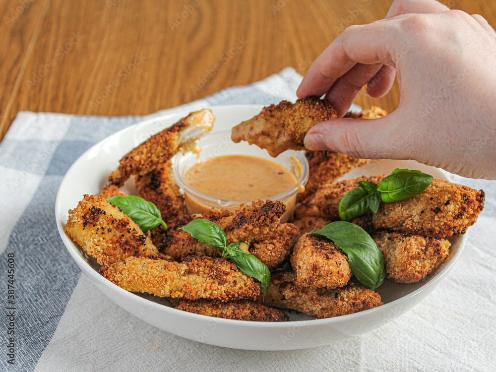 Hand holding chicken finger and dipping into the sauce. A plate of ...