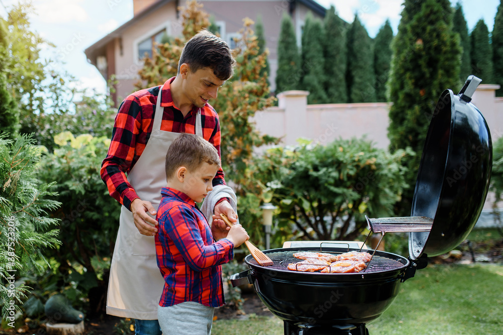 Young father and his son are cooking barbecue on an outdoor grill in ...