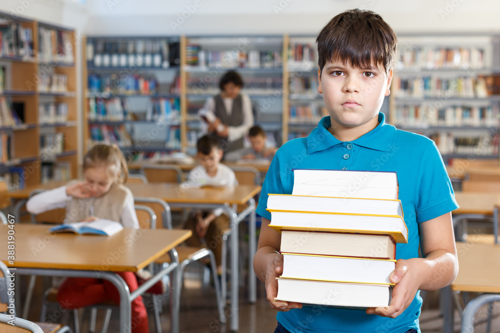 Upset tween boy standing in school library with pile of books in hands ...
