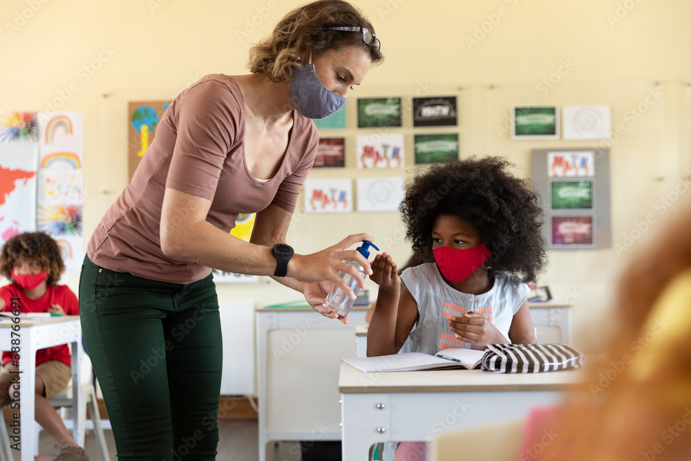 Female teacher wearing face mask sanitizing hands of girl in class ...