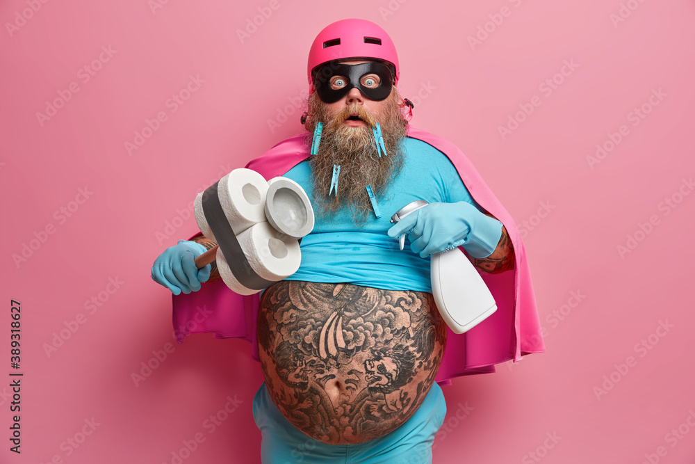 Stupefied bearded cleaning superhero holds plunger and detergent wears ...