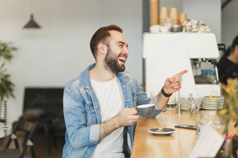 Laughing young man in denim jacket sit in coffee shop cafe hold cup of ...