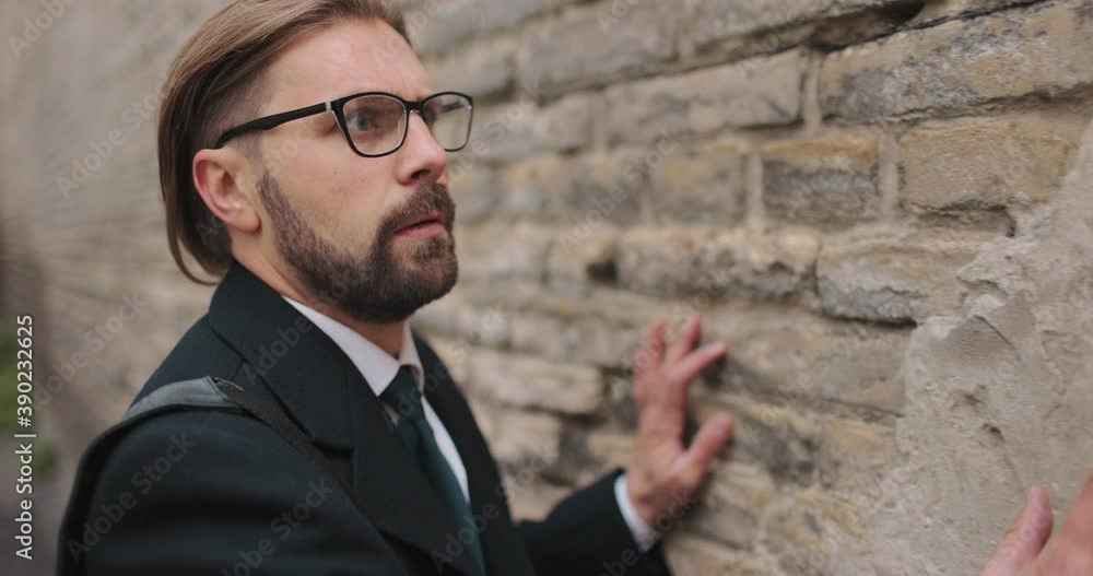Scared bearded man in formal suit and eyeglasses creeping near brick ...