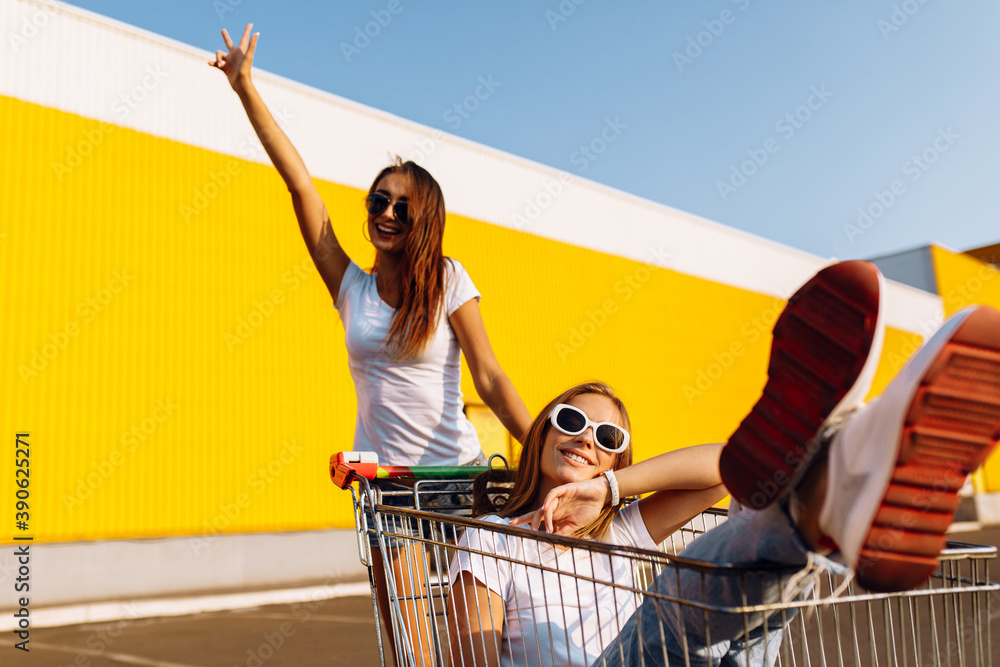 Excited young women friends ride shopping cart in city against yellow ...