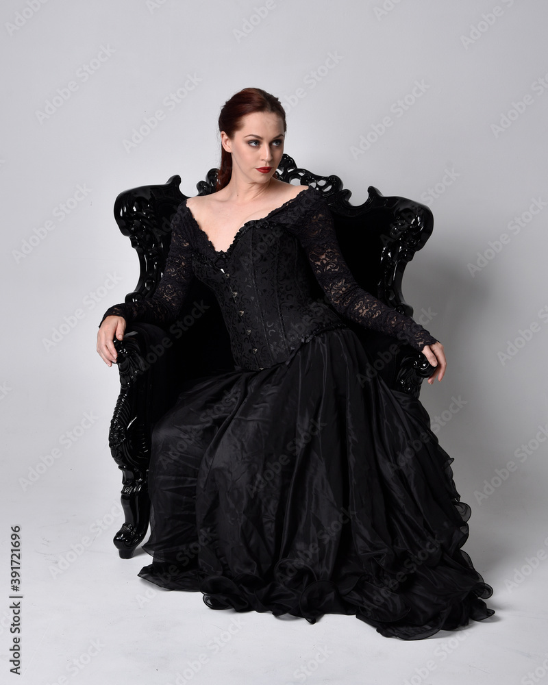 full length portrait of woman wearing black gothic dress, sitting on a ...