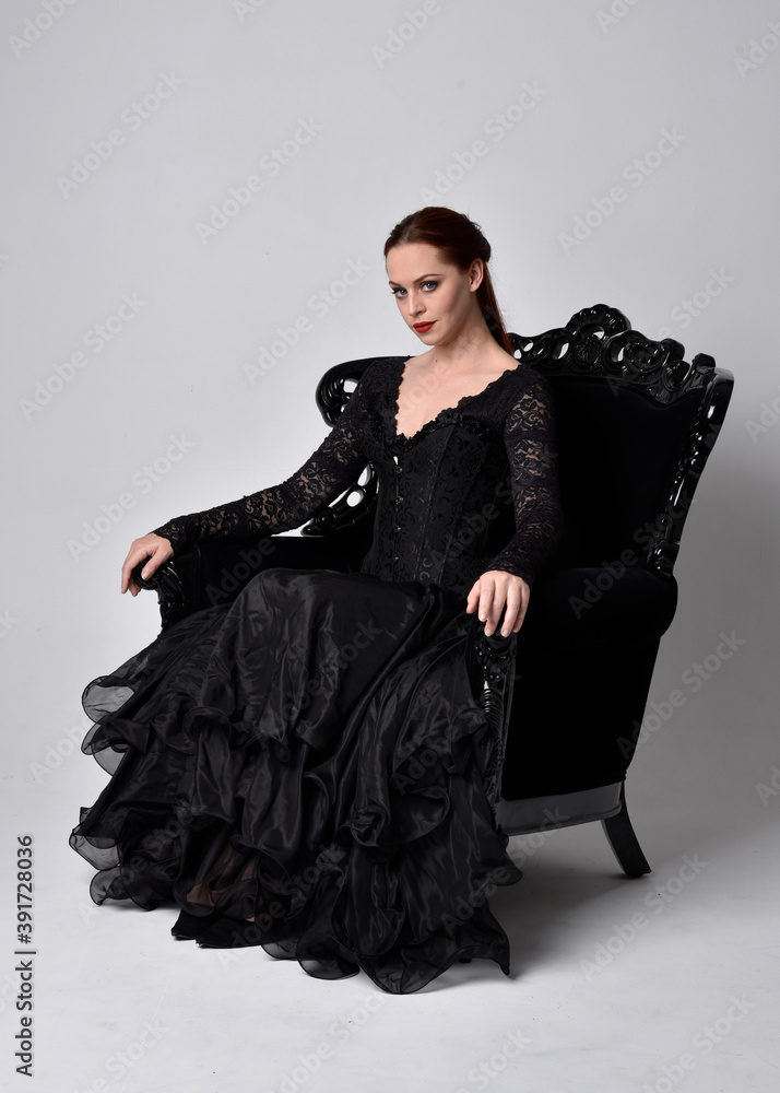 full length portrait of woman wearing black gothic dress, sitting on a ...