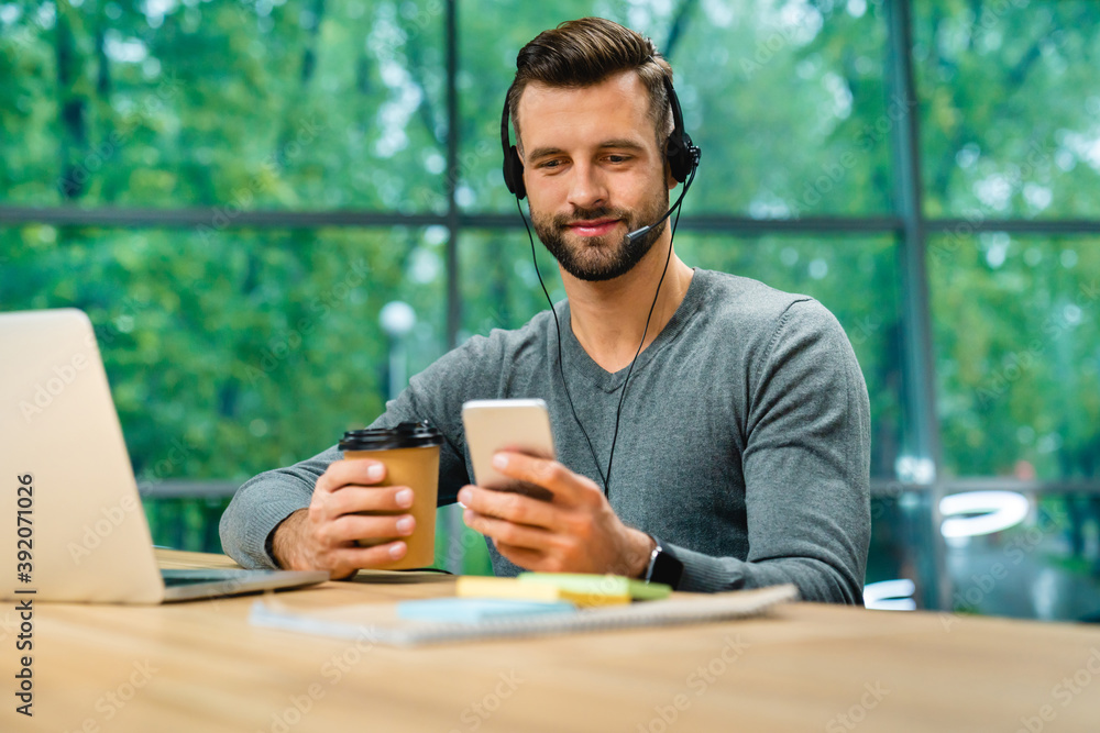 Smiling good-looking 30s man working with headset and phone at office ...