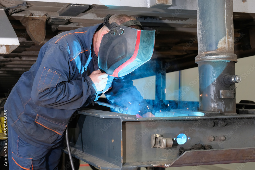 A welder in a helmet works in a garage, welding a cargo trailer ...