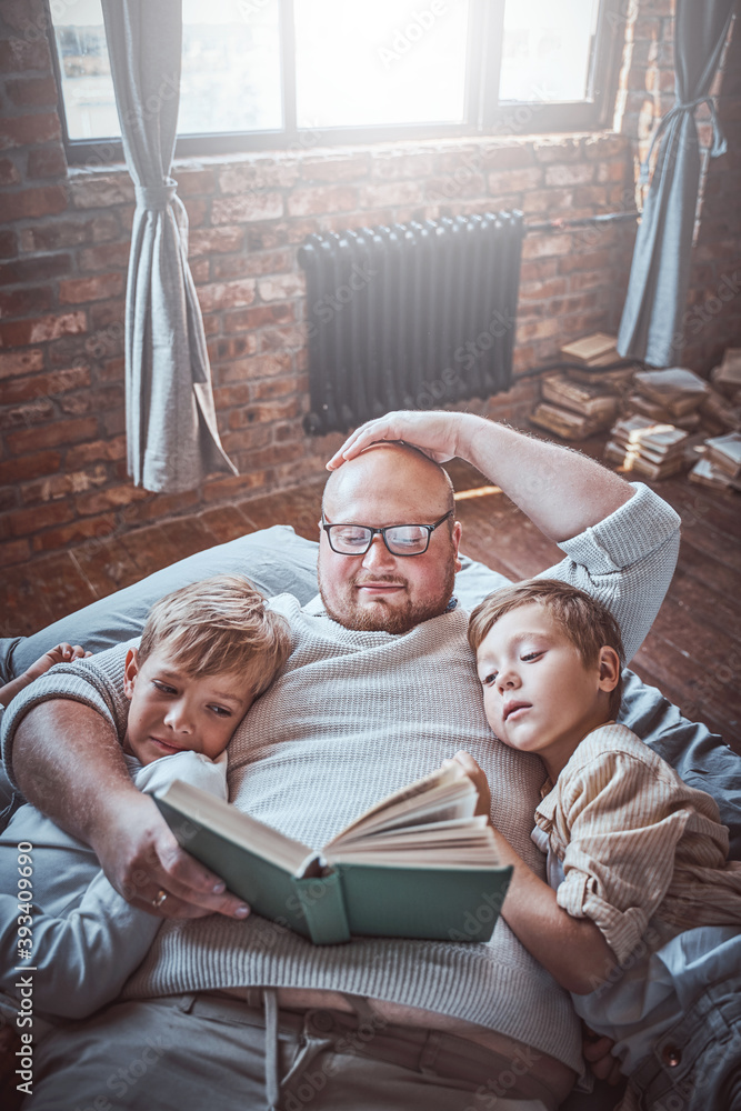 Two cheerful boys with their bald father reads a book lying on a ...