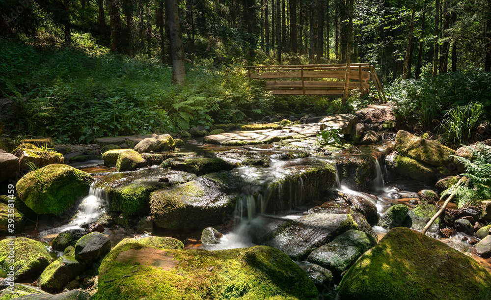 Idyllischer Bach im Wald mit großen bemoosten Steinen und Holzbrücke ...