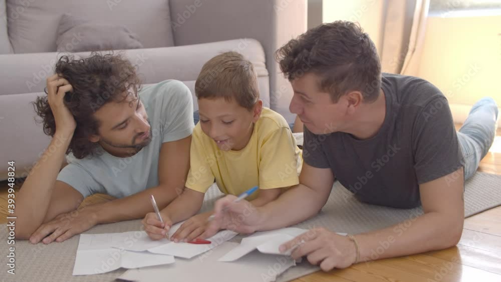 Two fathers helping focused boy with home task, lying on floor at home ...