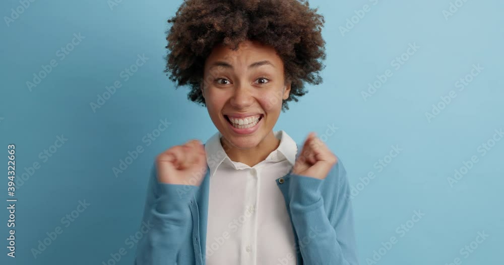 Overjoyed dark skinned woman cheers over something raises hands and ...