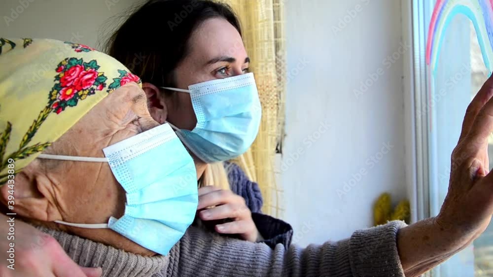 An old grandmother in a protective mask looks out the window with her ...