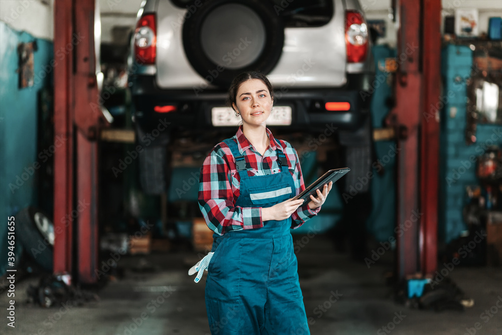 A young smiling mechanic female poses in a uniform with a tablet in her ...