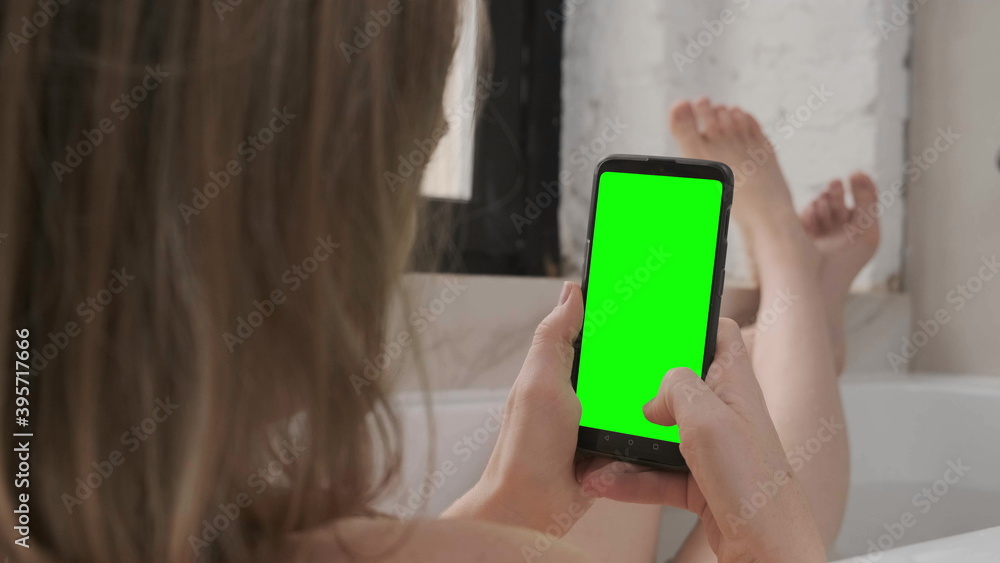Young naked woman lies in light white bathtub and looks at the phone with green screen. Lady ...