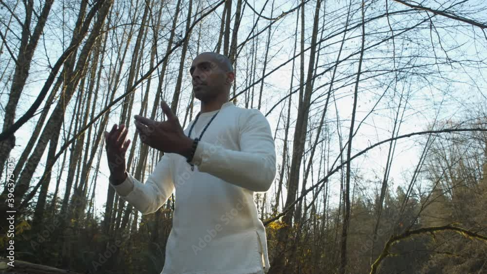 Indian male performs Tai Chi raising arms outside in a forest on a cold ...