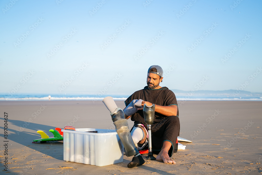 Pensive handsome surfer sitting on beach near surfboard and putting on ...