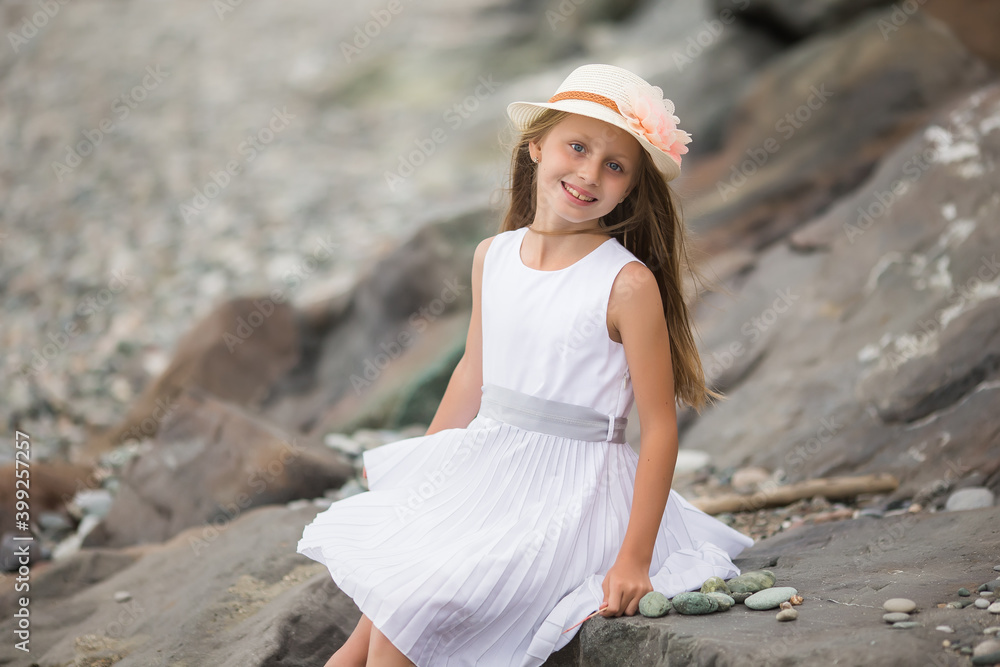 A cute smiling blonde girl 9 years old in a hat and white dress sits on ...