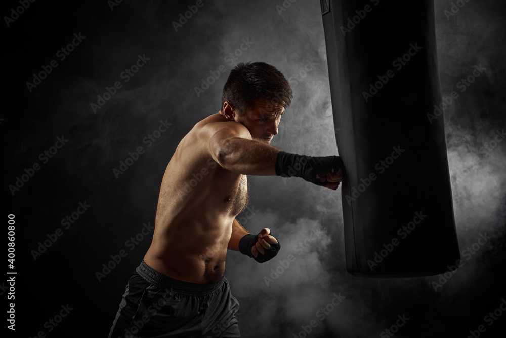 sporty shirtless boxer in black boxing wraps punching in boxing bag on ...