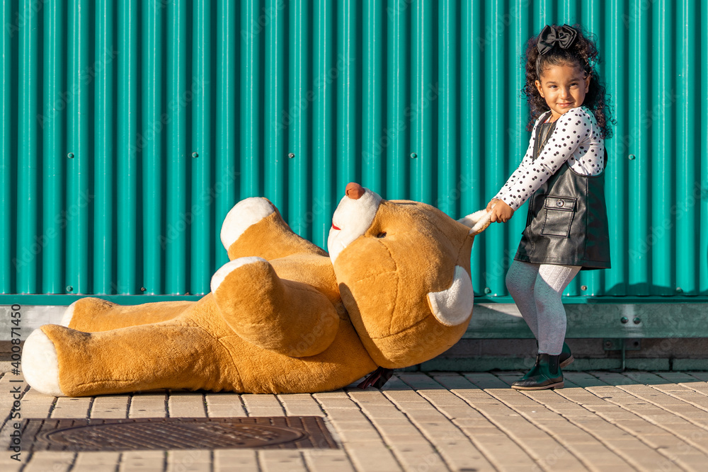 Girl dragging teddy bear on footpath while standing against green wall ...
