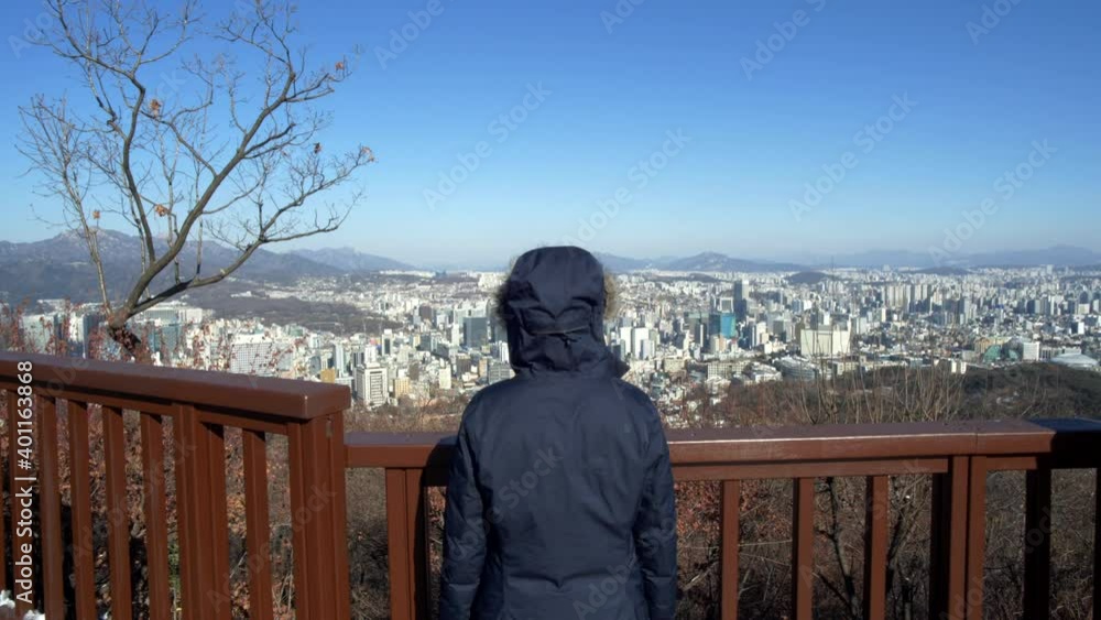 Hooded person with blue jacket looks out at the Seoul City skyline from ...