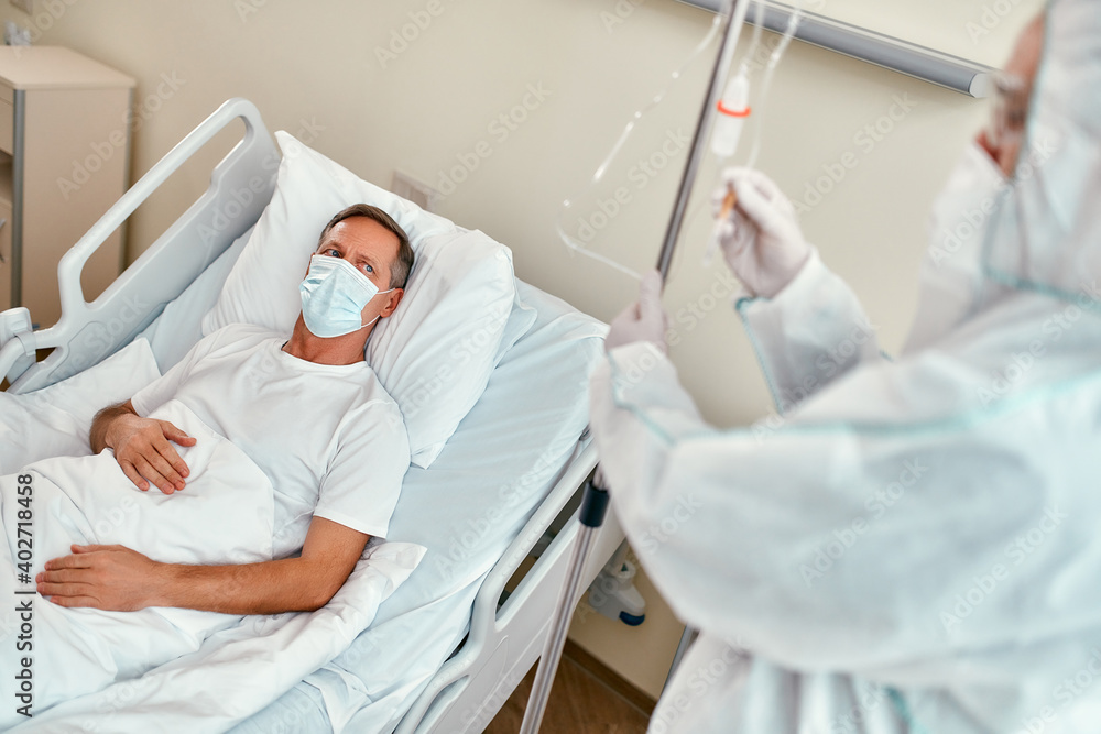A doctor in a protective suit, respirator and goggles puts an IV drip ...