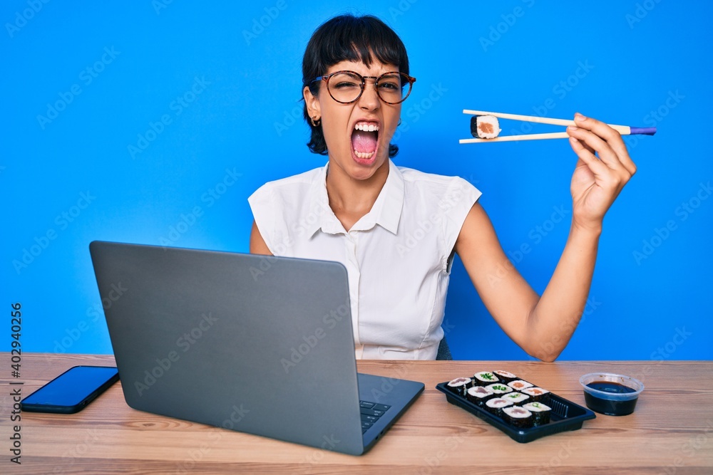 Beautiful brunettte woman working at the office eating sushi angry and ...