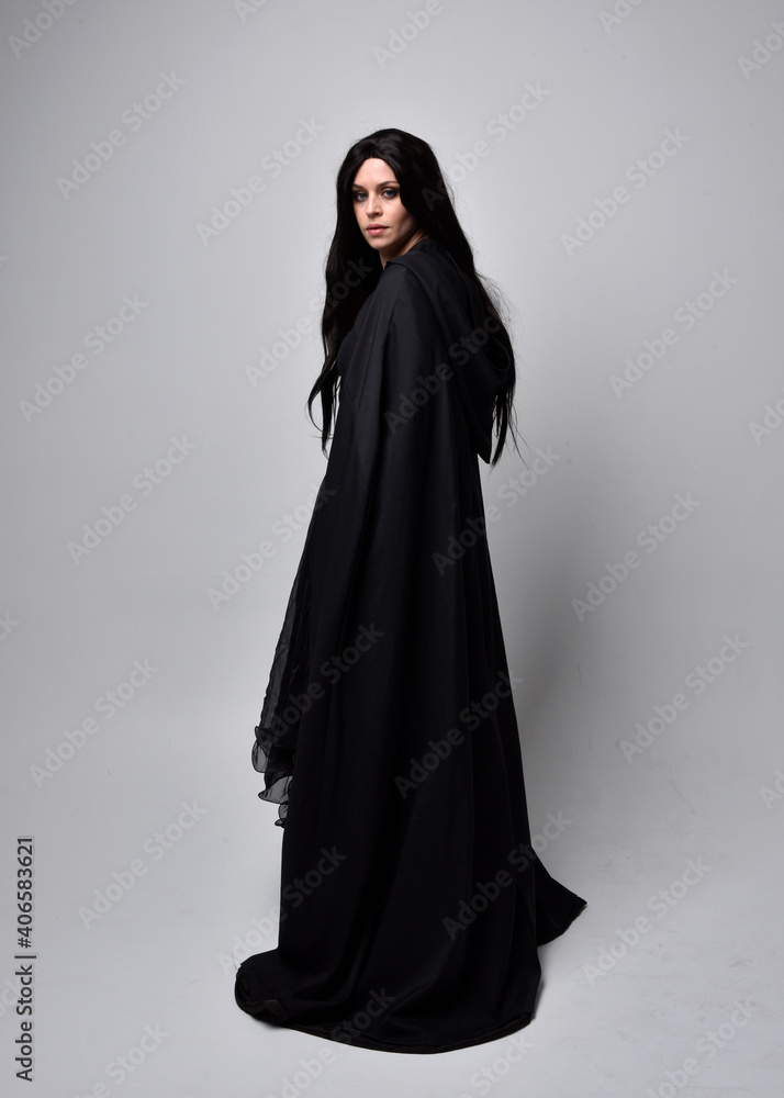 Full length portrait of pretty black haired woman wearing long dark gown nada cloak. Standing ...
