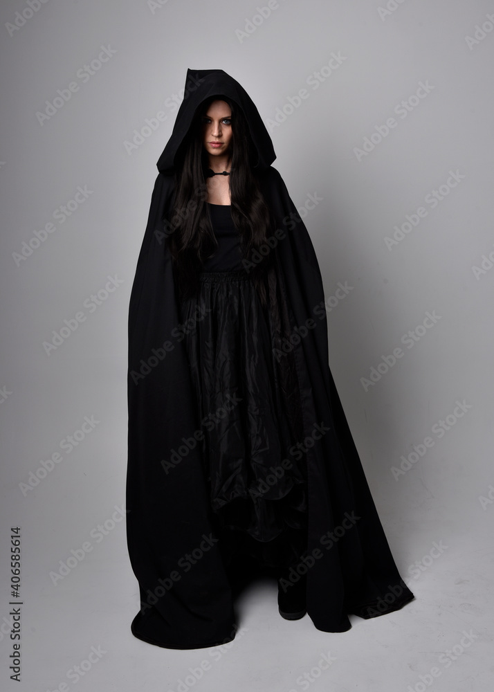 Full length portrait of pretty black haired woman wearing long dark gown and a cloak. Standing ...
