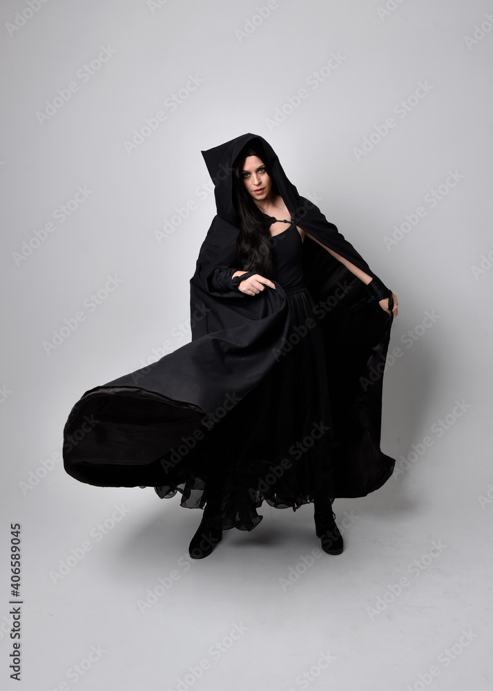 Full length portrait of pretty black haired woman wearing long dark gown and a cloak. Standing ...