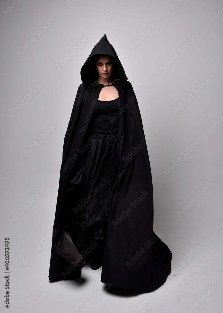 Full length portrait of pretty black haired woman wearing long dark gown and a cloak. Standing ...