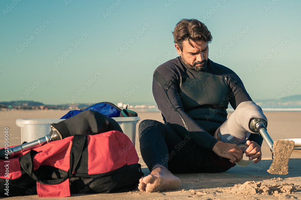 Focused male surfer wearing wetsuit for surfing, sitting on ocean beach ...