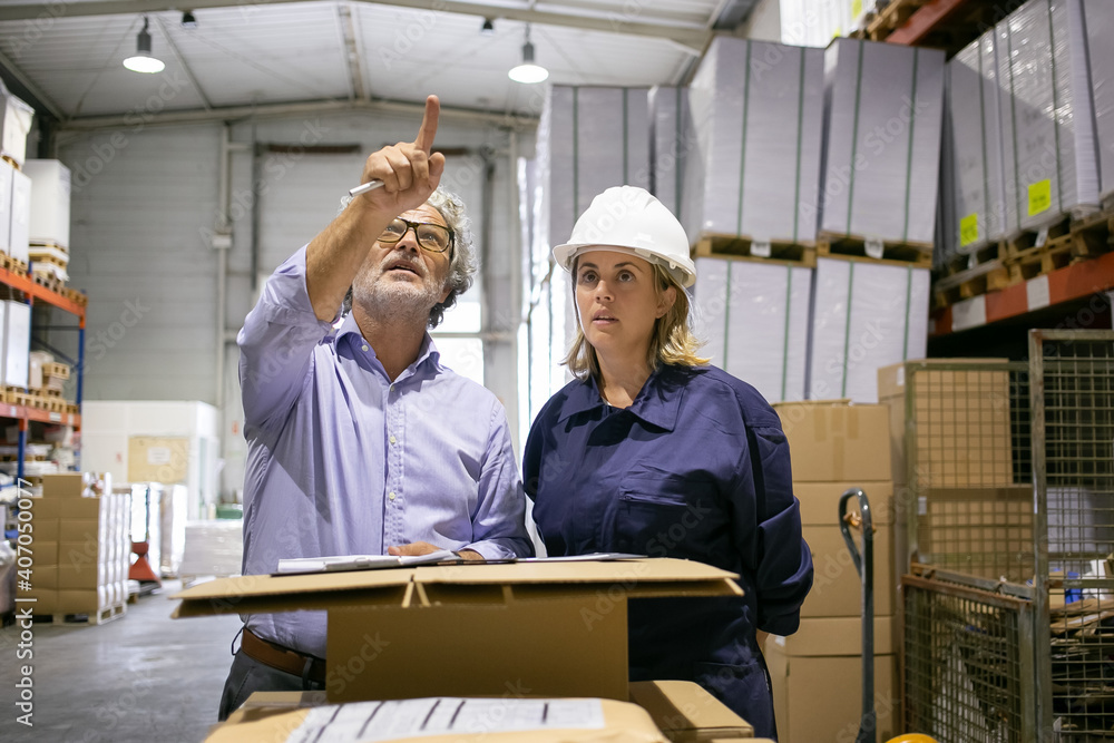 Mature inspector checking warehouse, pointing up and consulting female ...
