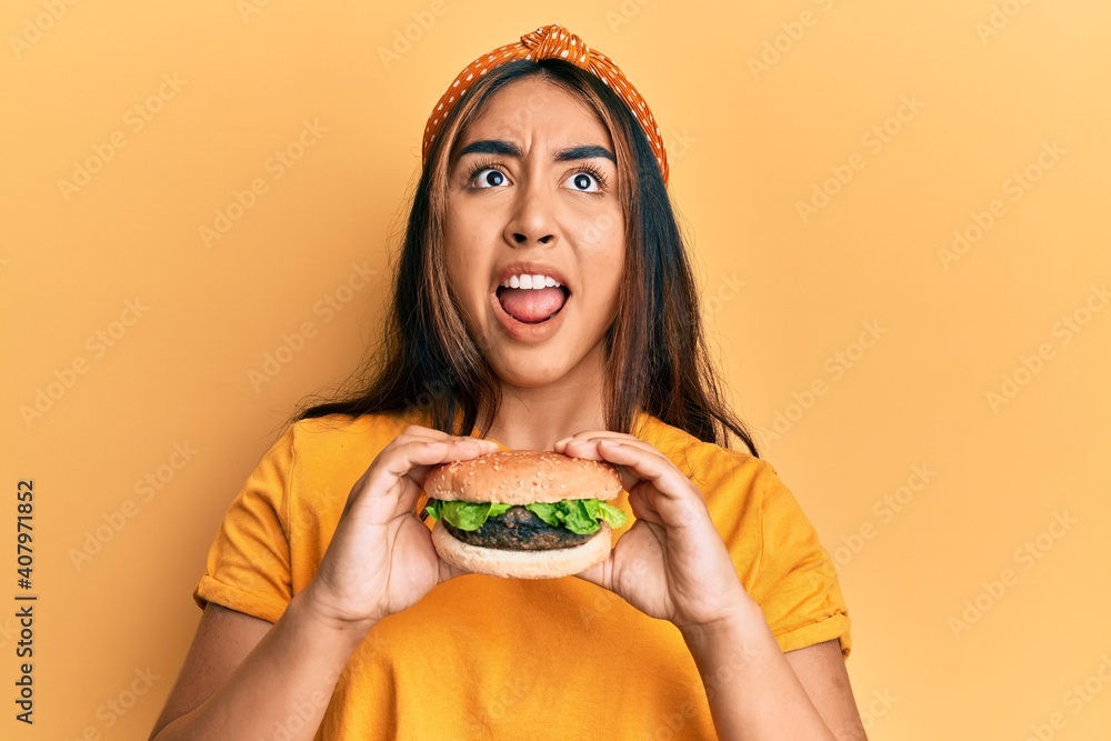 Young latin woman eating a tasty classic burger angry and mad screaming ...