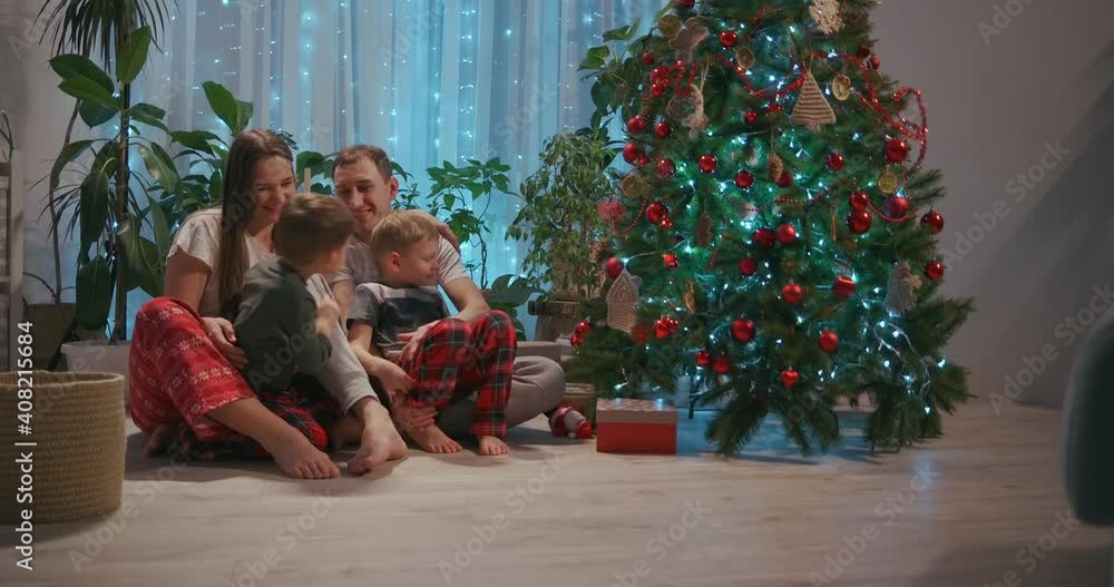 A family with two children sit together at the Christmas tree and ...