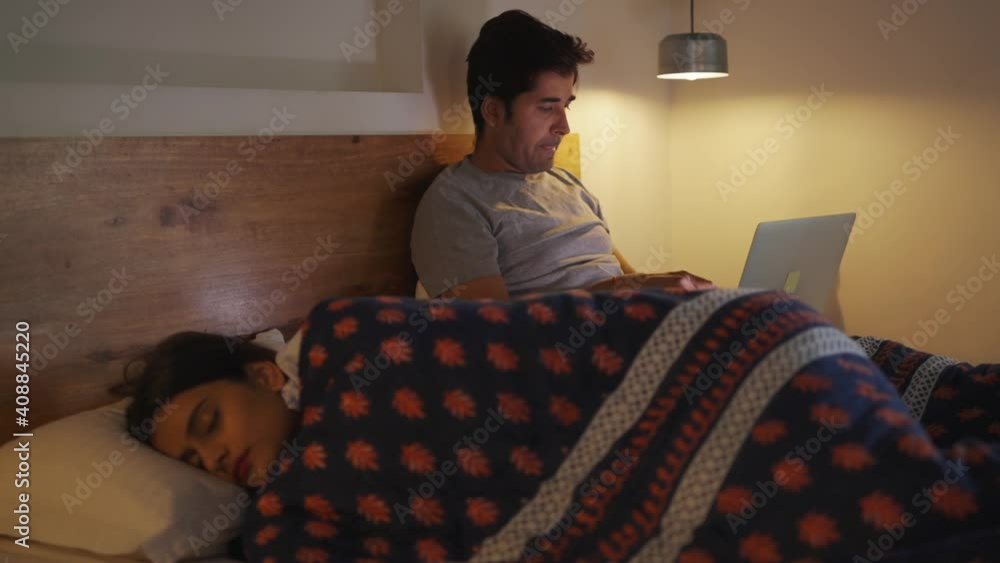 An overworked and workaholic young husband is sitting on a bed busy ...