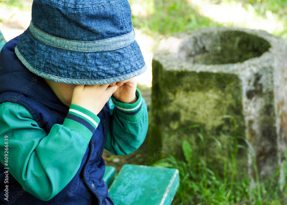 a little boy in a blue vest and a Panama hat is crying in a park in the ...