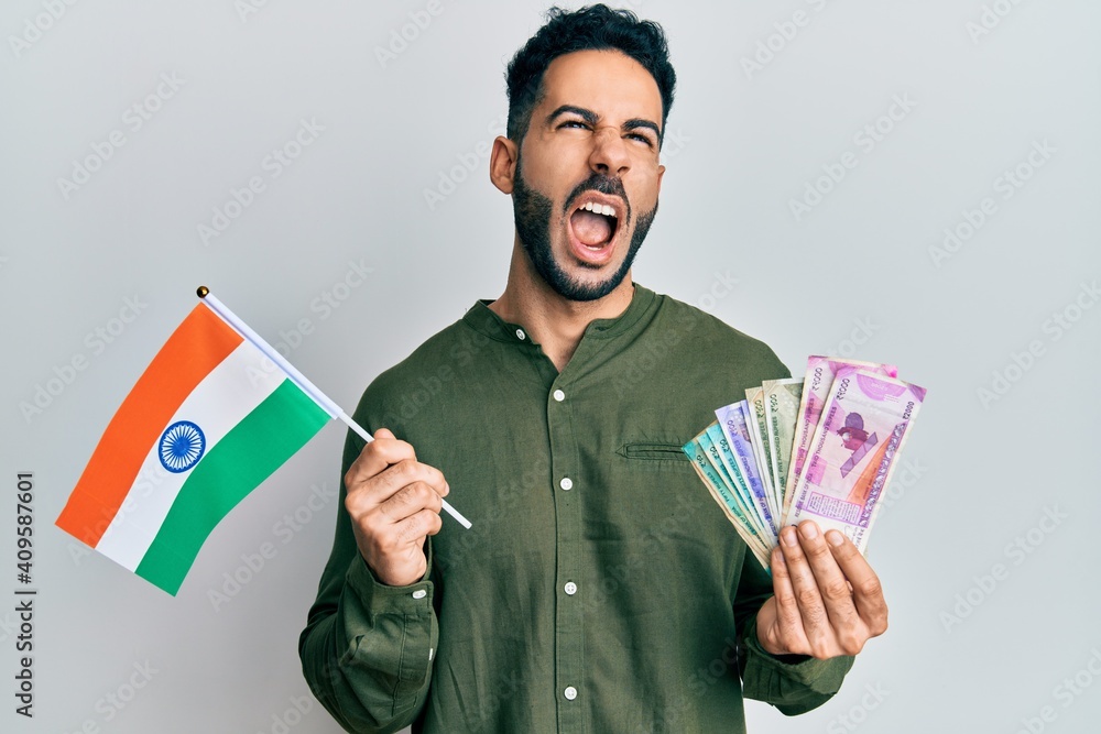 Young hispanic man holding india flag and rupee banknotes angry and mad ...