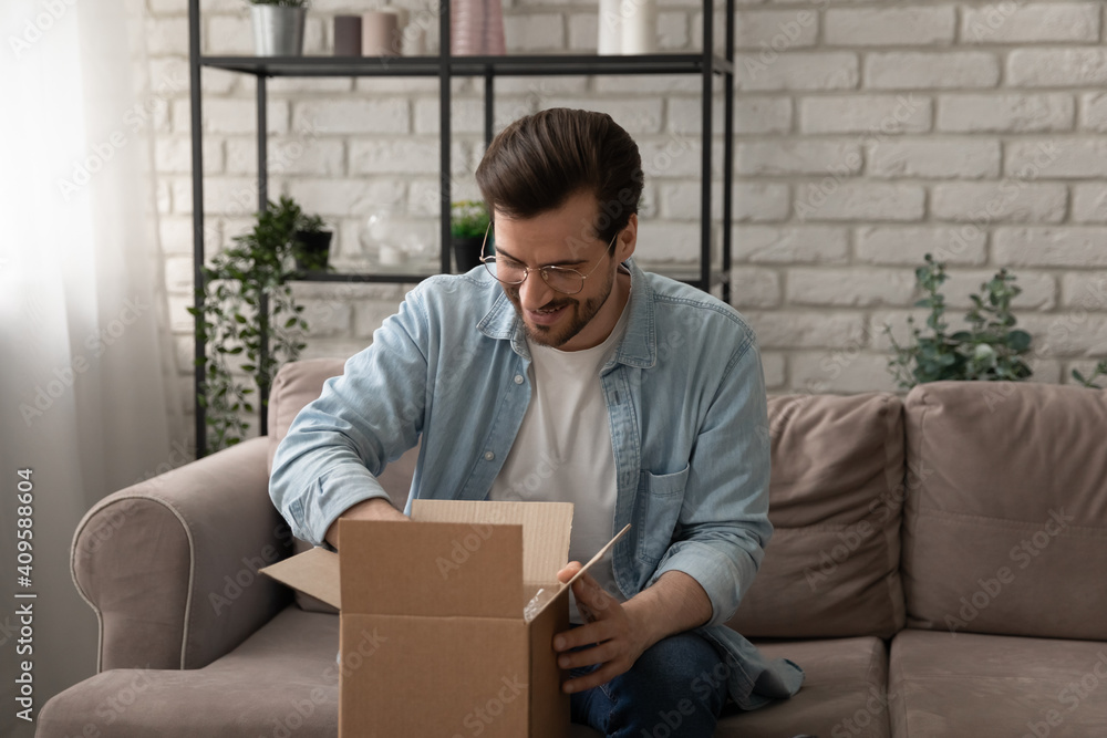 Curious young man wearing glasses unpacking awaited parcel, sitting on ...
