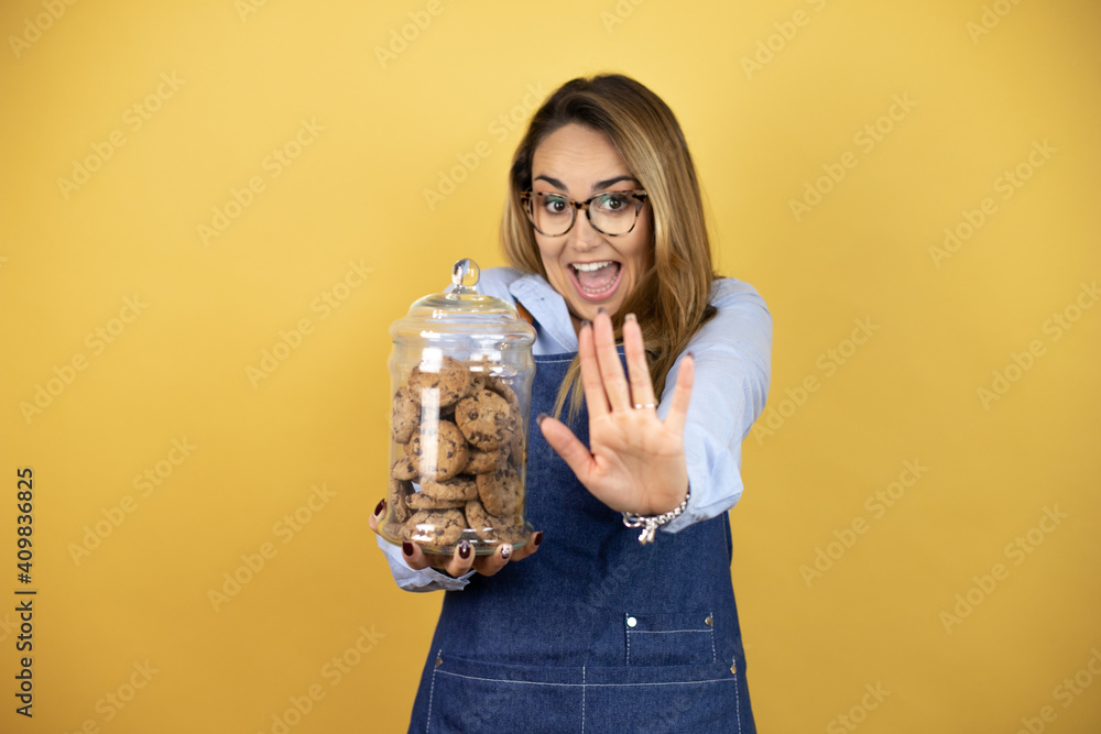 Young hispanic woman wearing baker uniform holding a cookies jar afraid ...