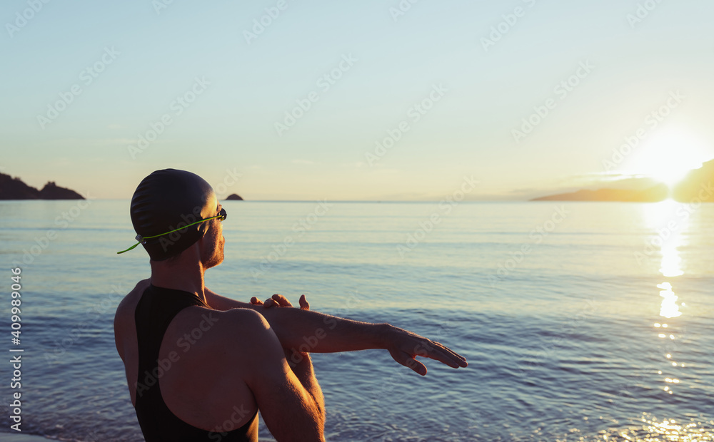 Back view of fit male swimmer in swimsuit and cap standing on seashore ...