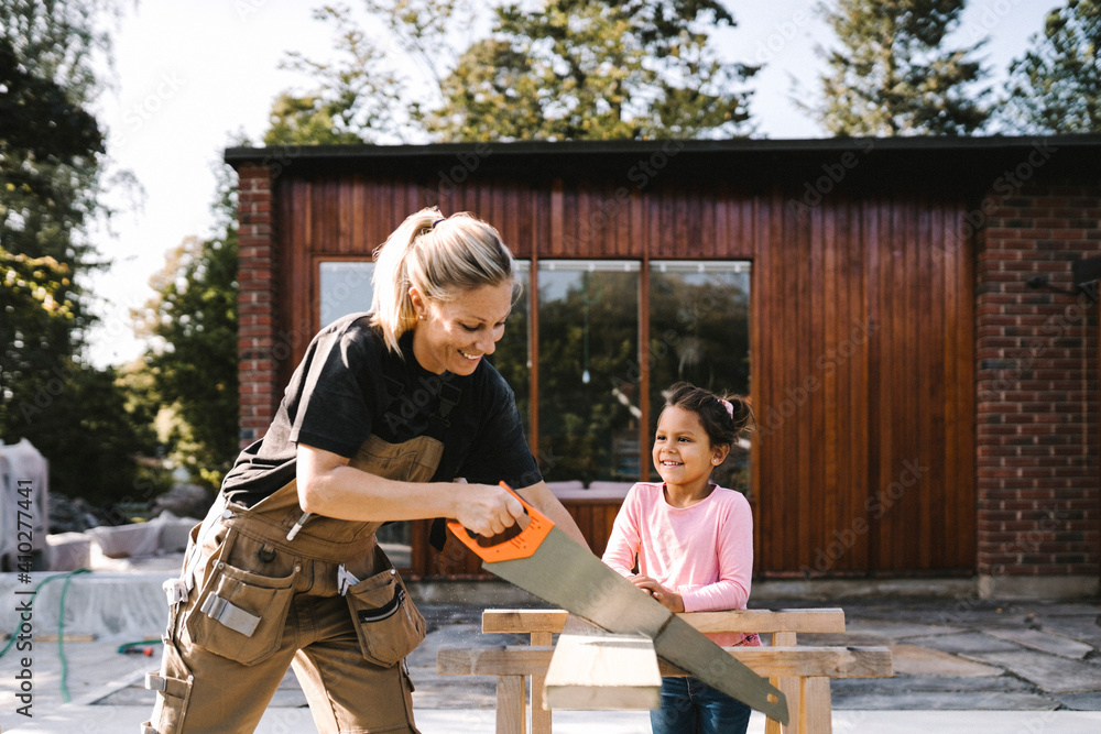 Smiling daughter looking at mother cutting plank with hand saw against ...
