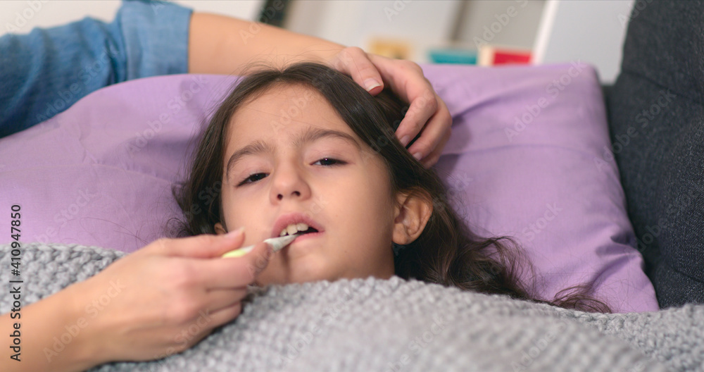 Young mother taking care of sick child lying in bed with flu is measuring temperature using ...