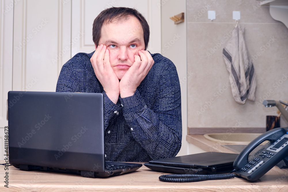 Sad man sits in the kitchen at the laptop in the home office. Workplace ...