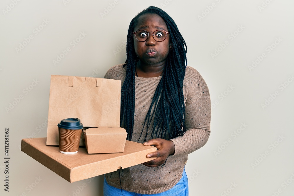 Young black woman with braids holding take away food puffing cheeks ...