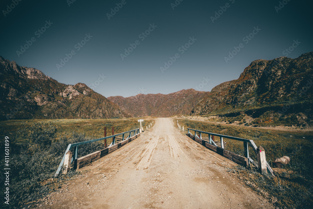 Wide-angle mountain scenery with an old wooden bridge and an earth road ...