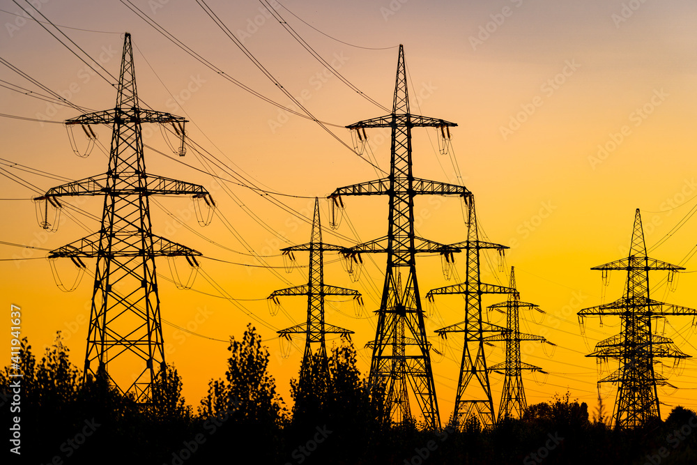 Power pylons reache into the sunset sky. Silhouettes of big trees under ...