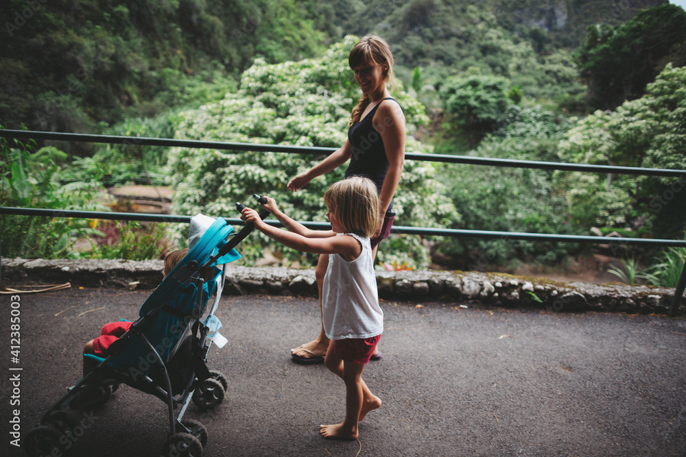 Cute young girl helping push stroller on nature path - mom watching ...