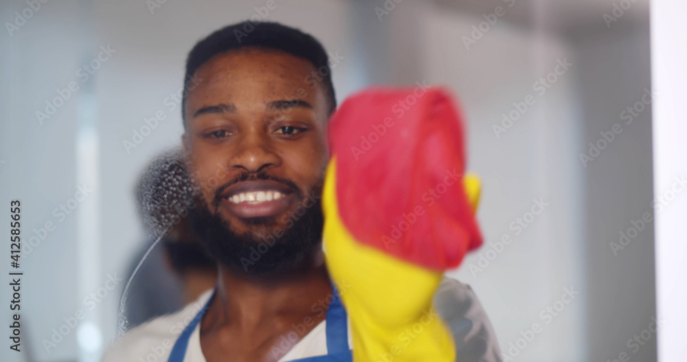 African male janitor using sprayer and rag to clean window in office ...